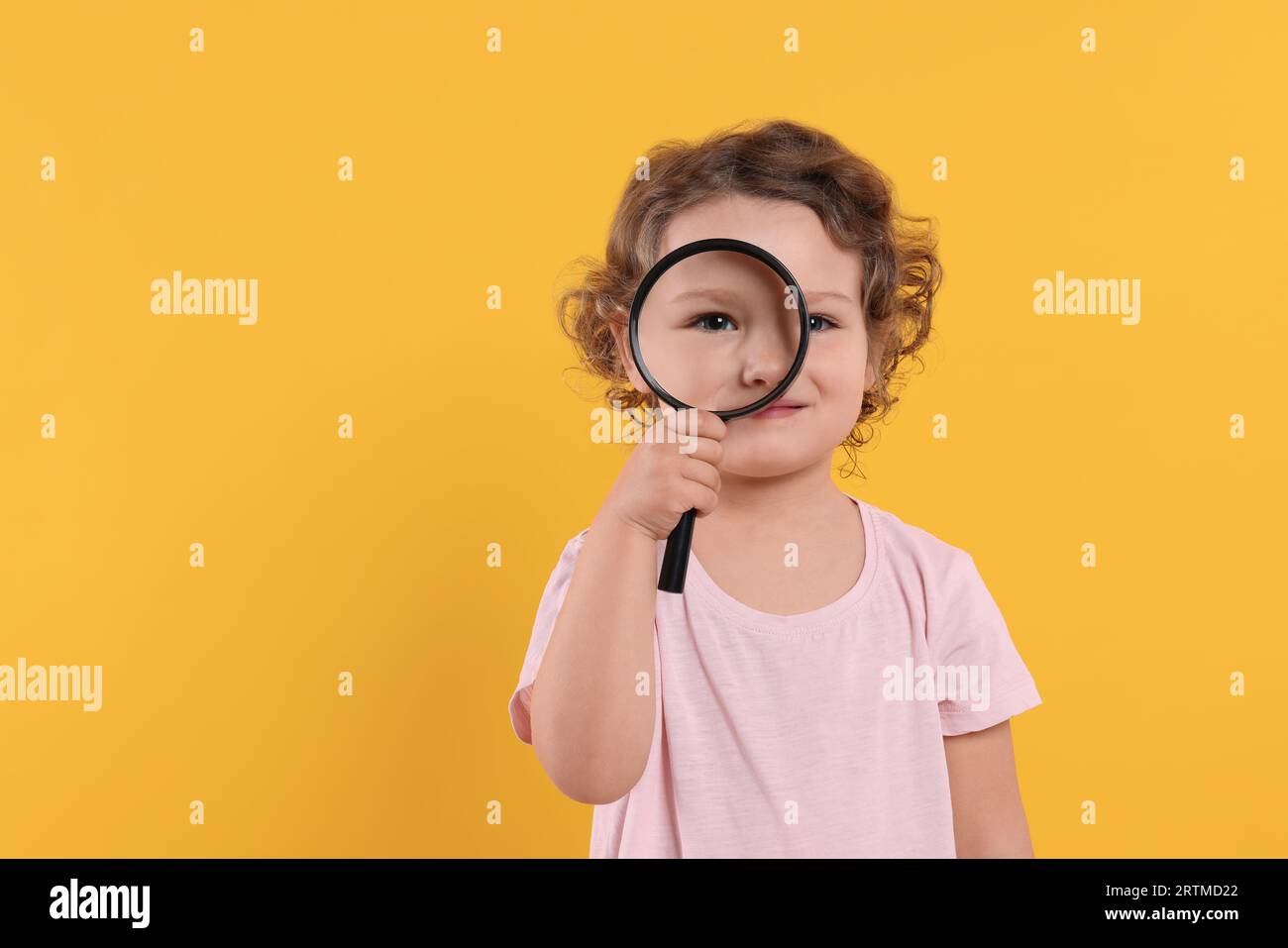 Cute little girl looking through magnifier glass on orange background, space for text. Searching ...