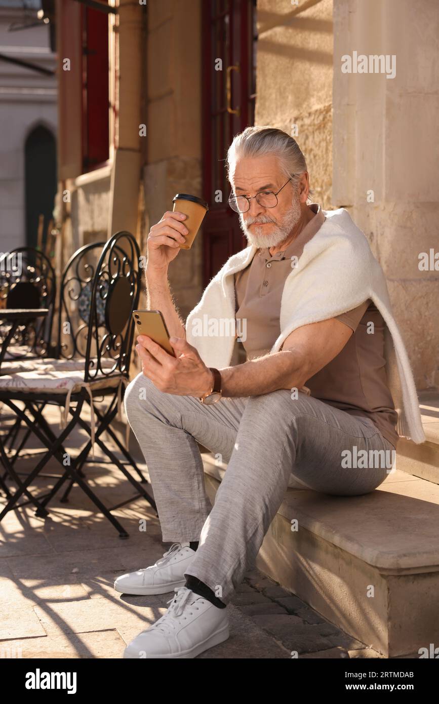 Handsome senior man sitting on doorstep, using smartphone and drinking ...