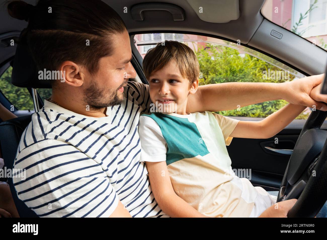 Father teaching his young son how to drive car on his lap, looking at ...