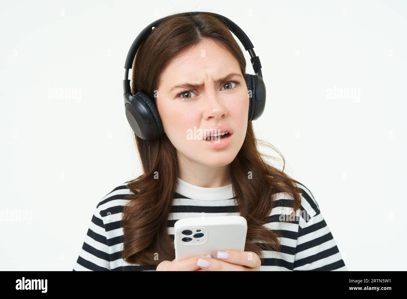 Portrait of girl in headphones with smartphone, looks confused at camera, frowns with perplexed ...