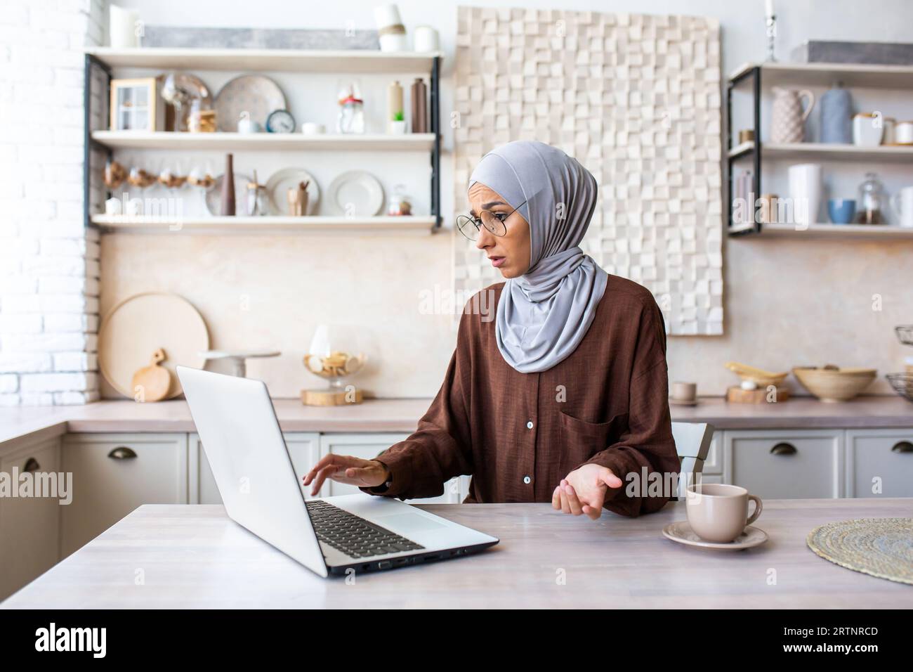 Young Muslim woman in hijab sitting worried in kitchen and using laptop ...