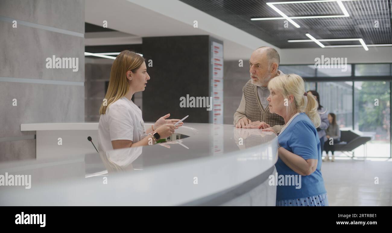 Elderly couple stand near reception desk in clinic lobby area and make ...