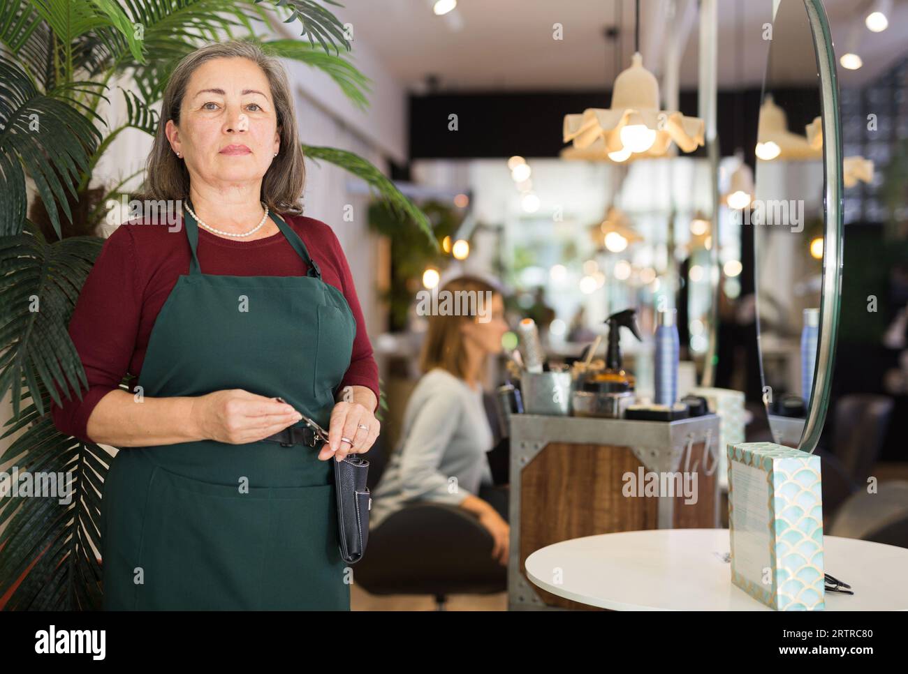 Experienced aged woman hairdresser standing in barbershop with scissors ...