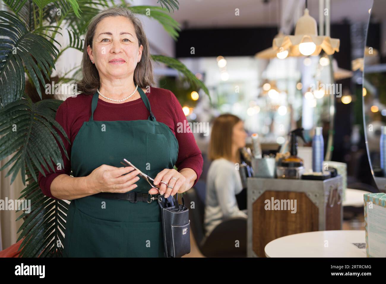 Smiling successful elderly female hairdresser posing in barber shop ...