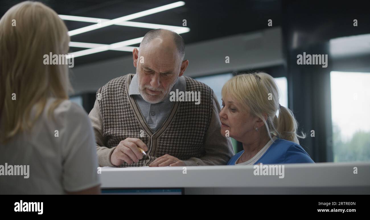 Elderly couple stand near reception desk in clinic lobby area, fill out ...