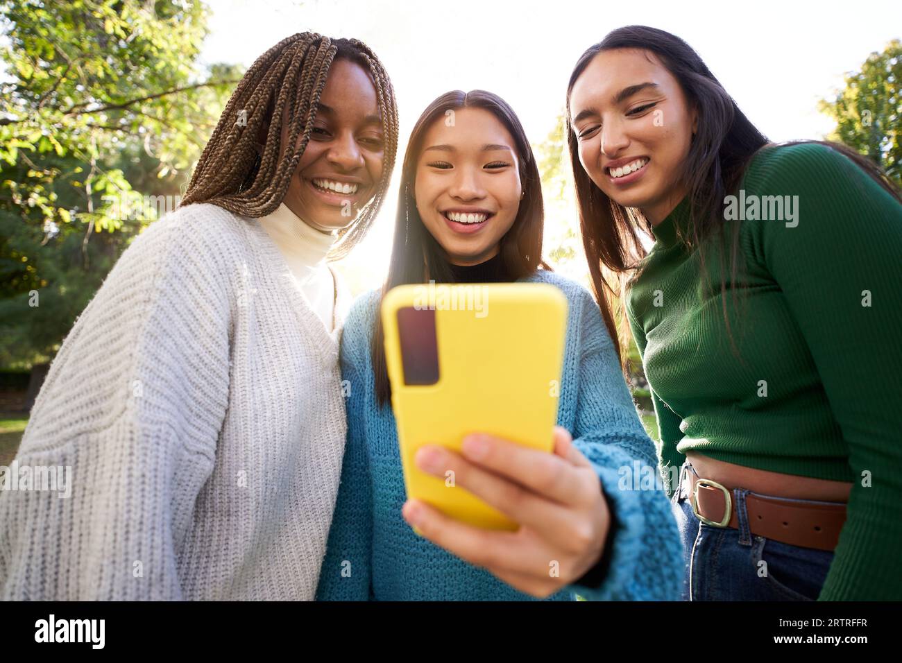 Three smiling multiracial young girls using cell phone outdoor park ...