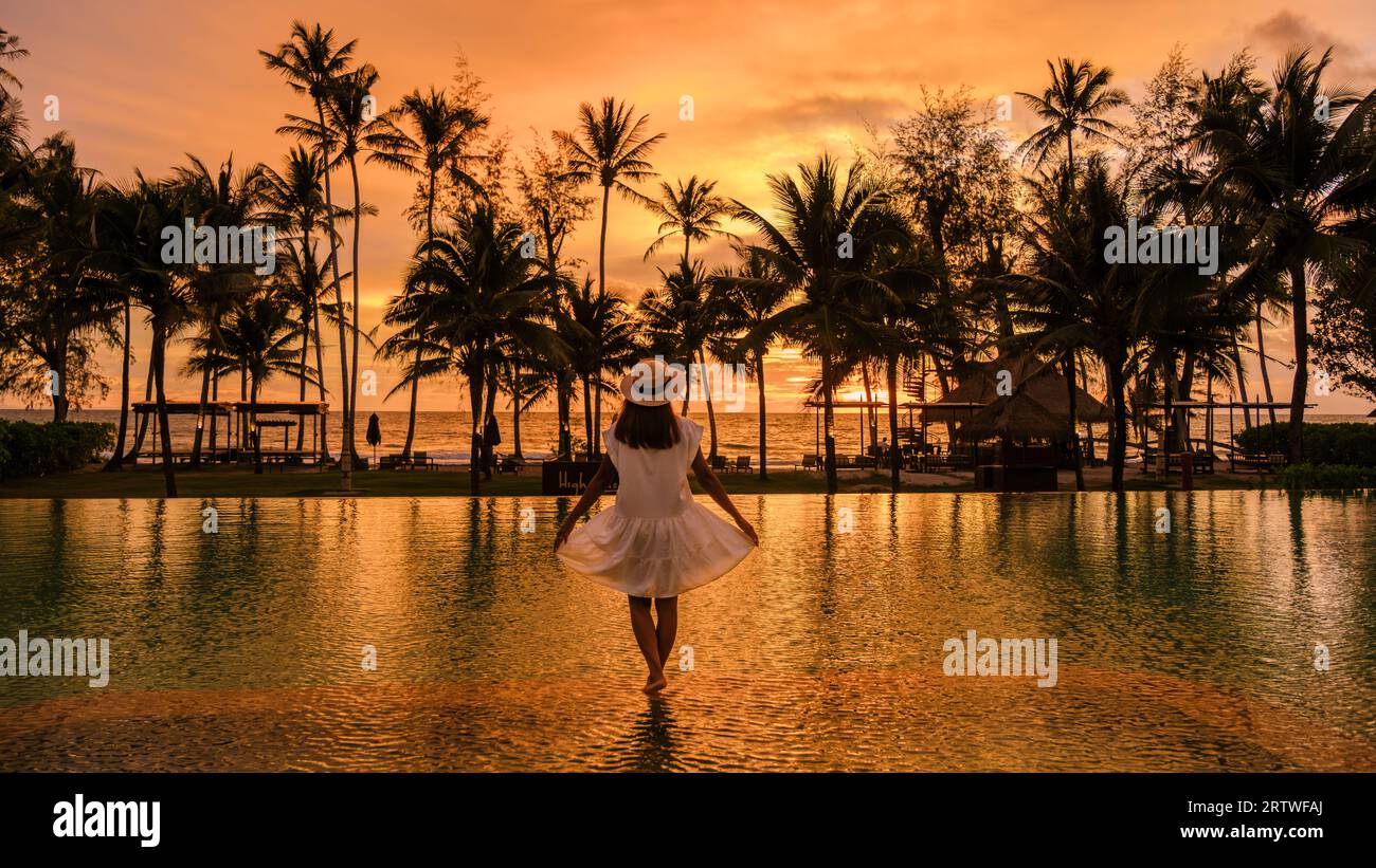 Asian woman watching sunset at a tropical swimming pool with palm trees ...