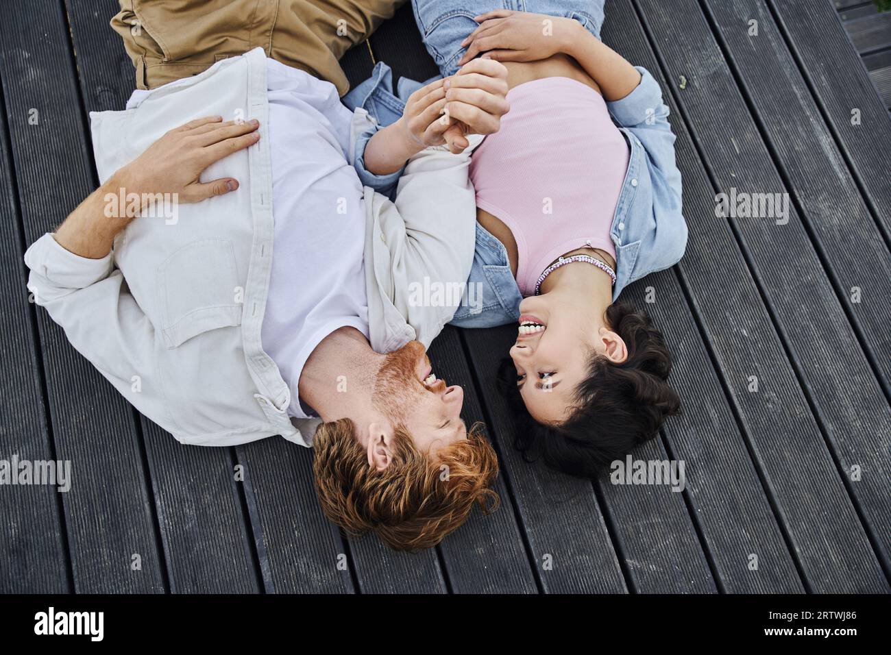 joyful interracial couple lying down on wooden porch and looking at ...