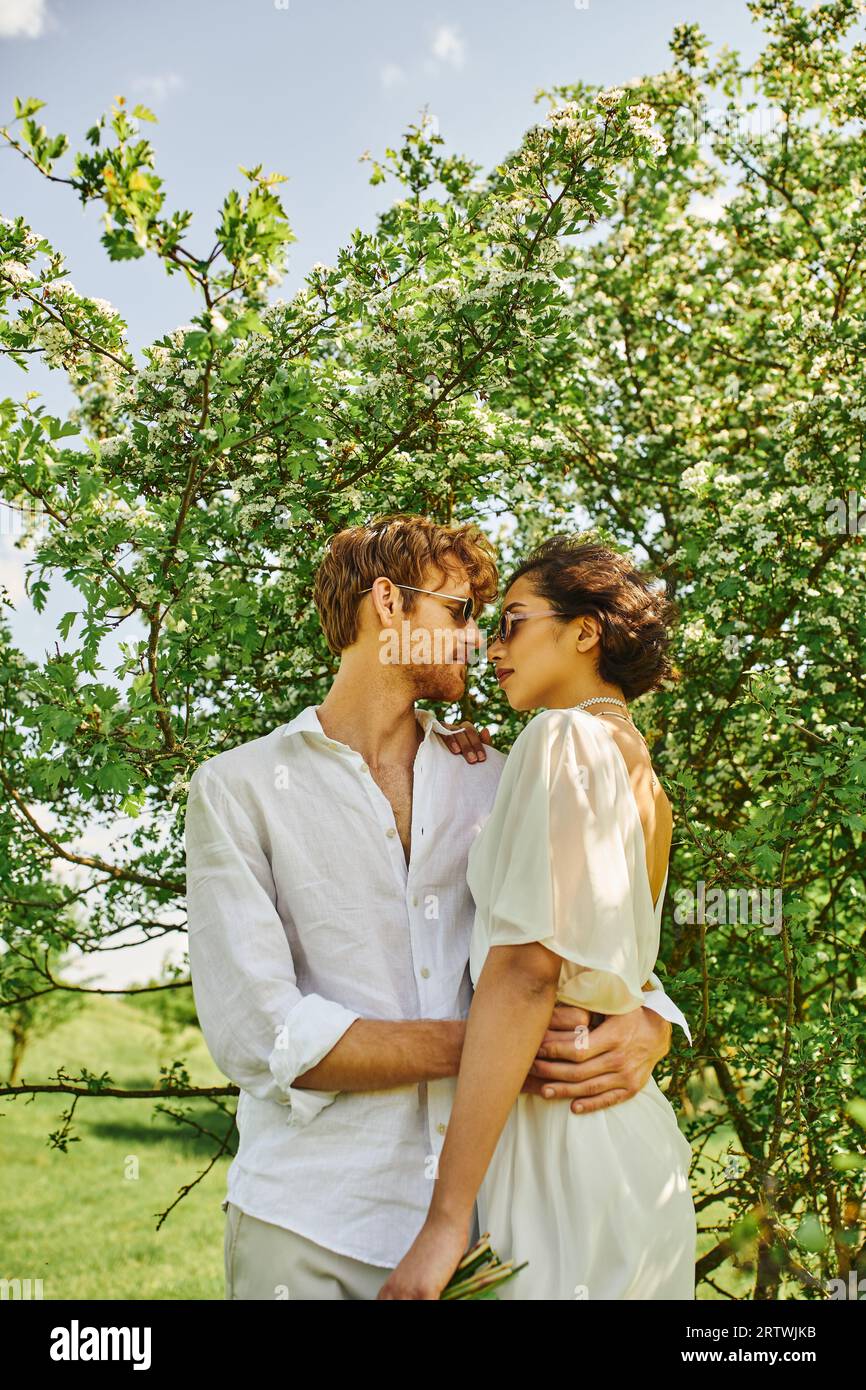 happy multiethnic newlyweds in sunglasses and wedding gown hugging in ...