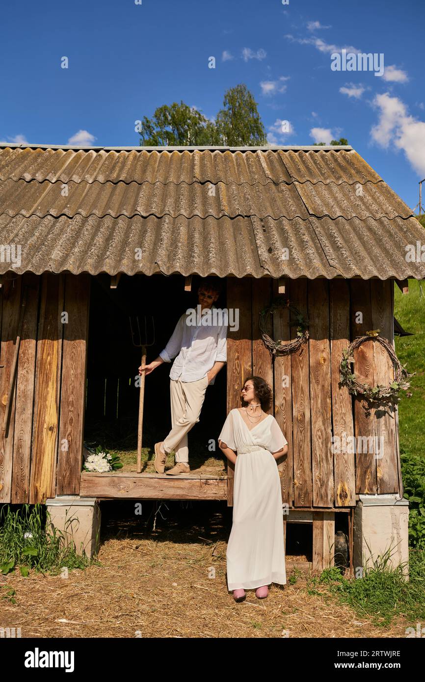 rustic wedding concept, interracial newlyweds posing near wooden barn ...