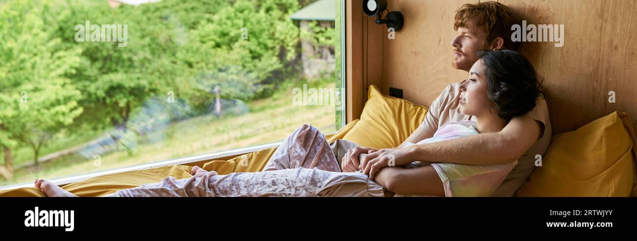 multicultural couple cuddling and enjoying view from window with green ...