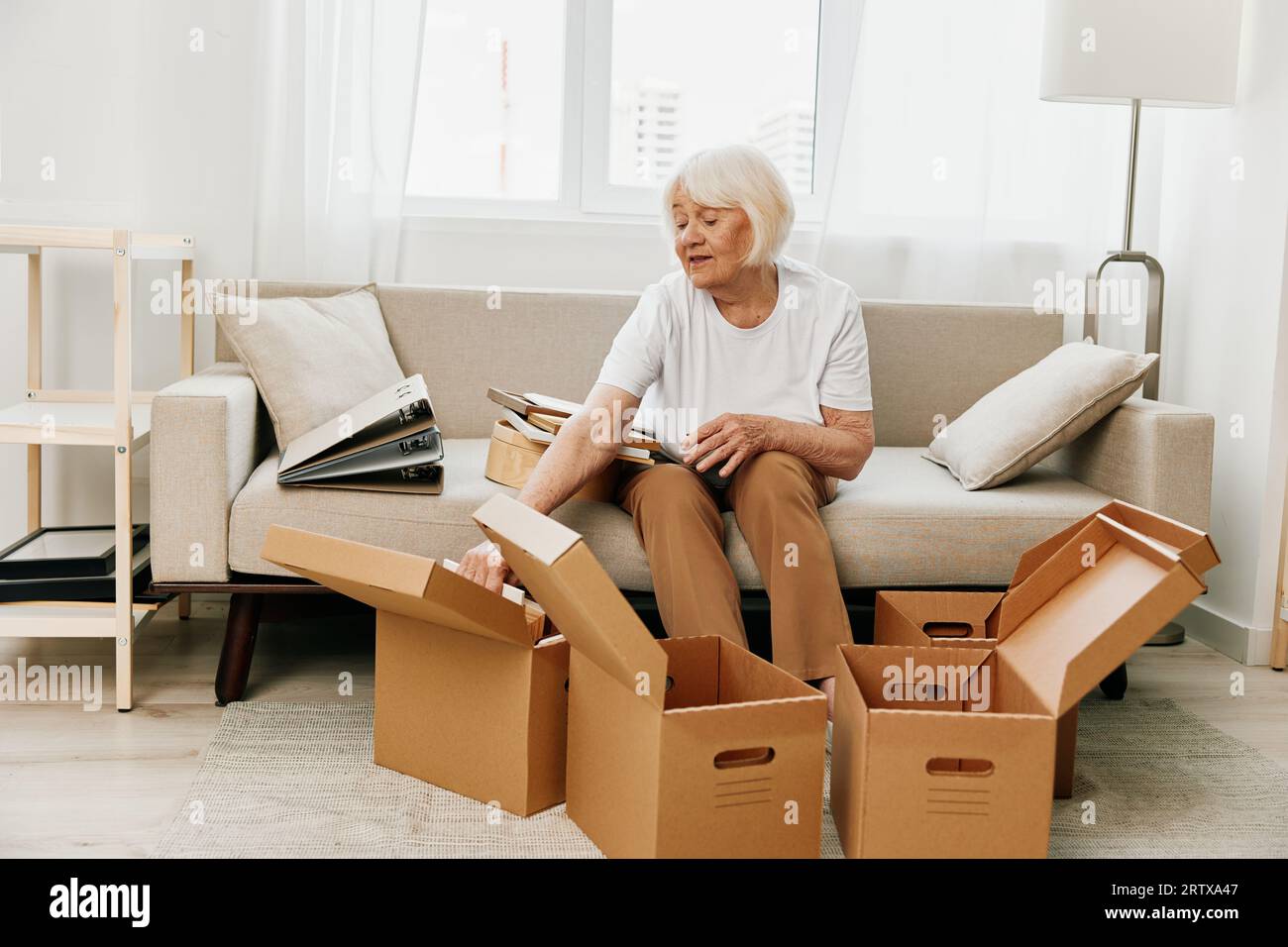 elderly woman sits on a sofa at home with boxes. collecting things with ...