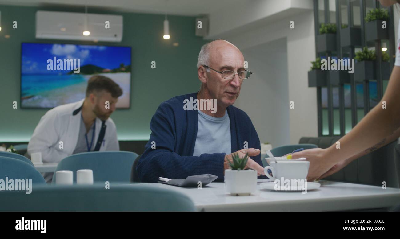 Elderly patient uses mobile phone in hospital cafeteria. Waitress ...
