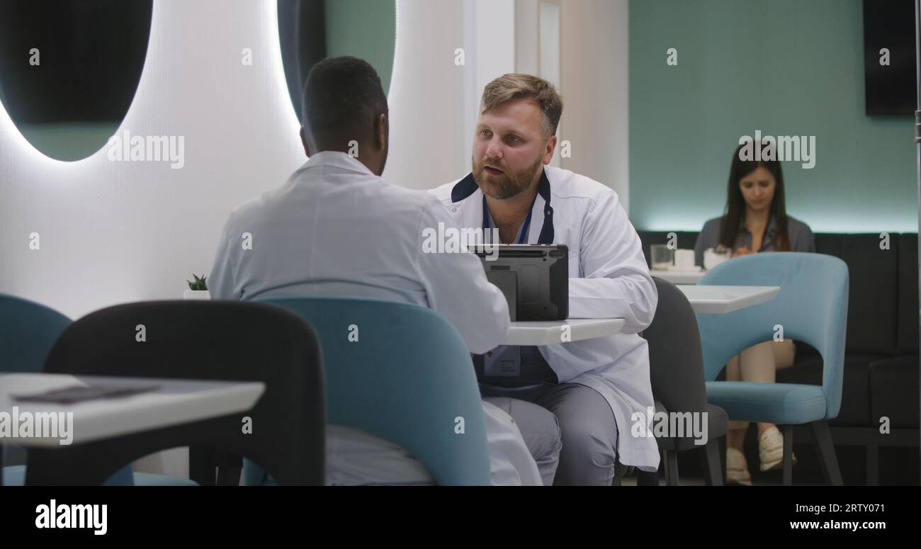 Two multicultural doctors sit in clinic cafe, discuss work. Male ...