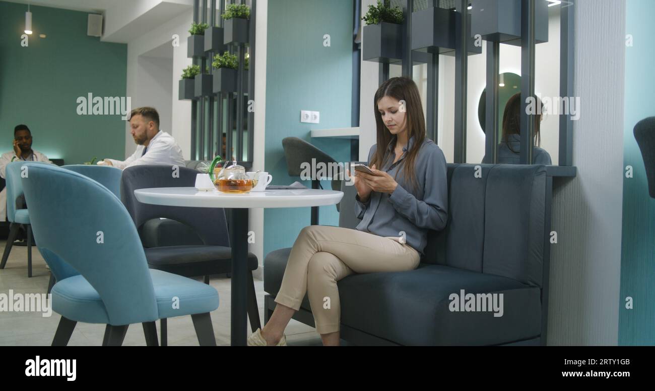 Adult woman sits on sofa in modern clinic cafe, surfs the Internet ...