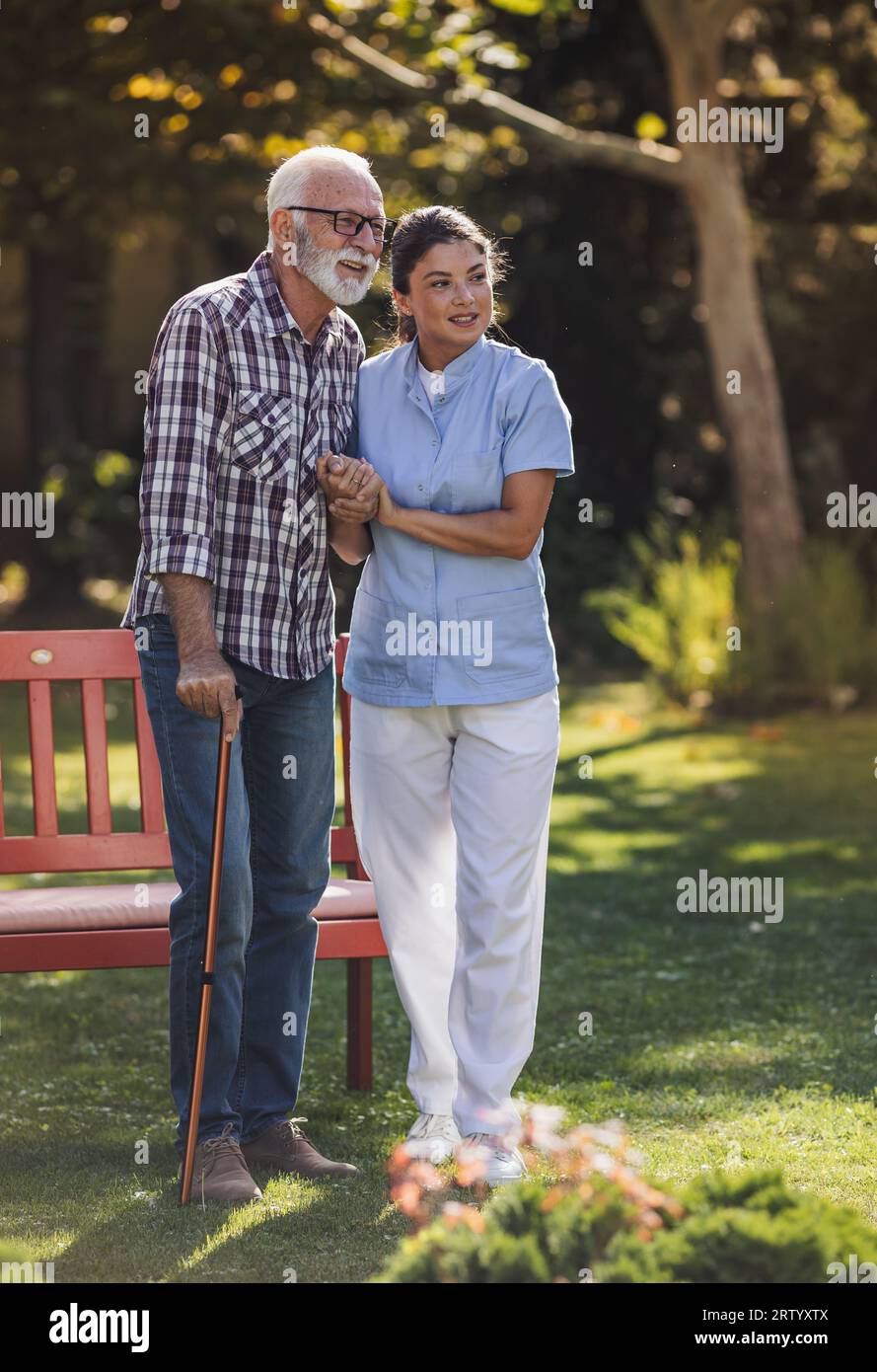 Young woman caregiver helping senior man to walk with stick in park ...