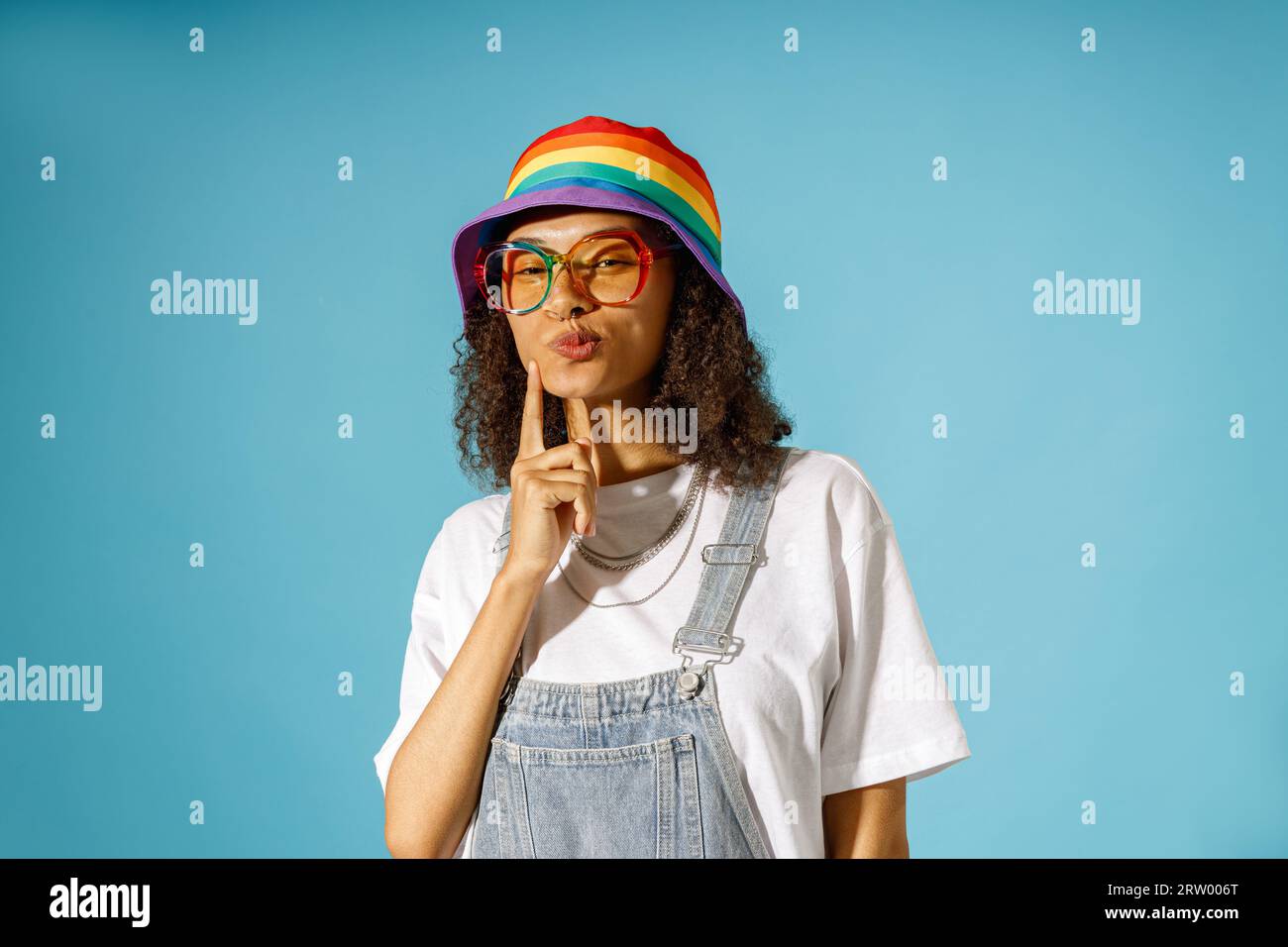 Woman in rainbow cap and eyeglasses touching cheek and looks camera over blue studio background ...