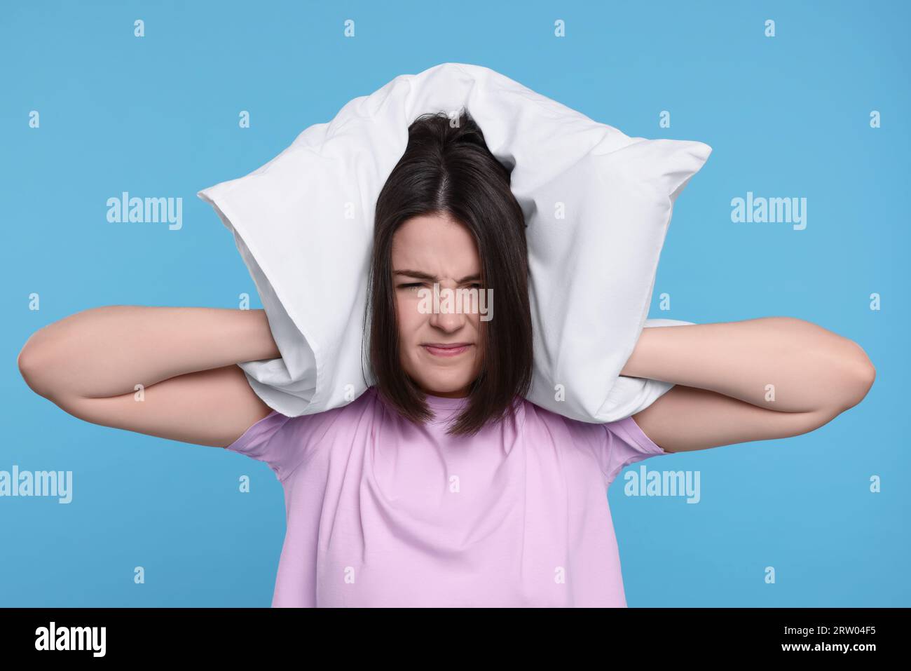 Irritated young woman covering ears with pillow on light blue ...