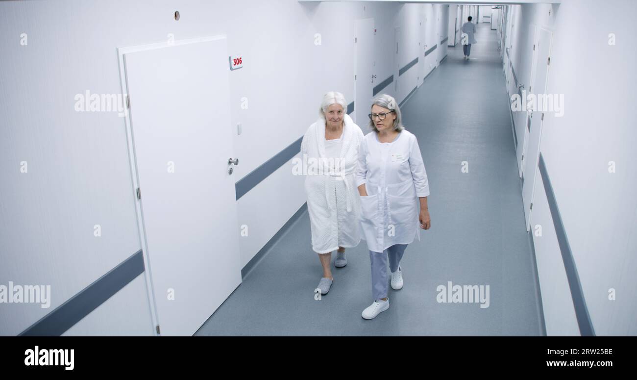 Bright clinic corridor: Female doctor and elderly woman walk down the ...