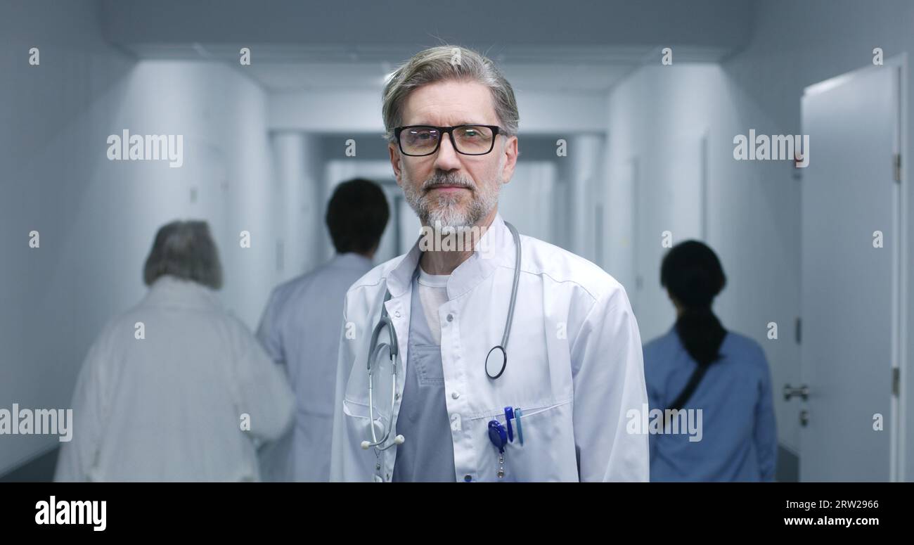 Mature doctor stands in modern clinic corridor. Professional medic puts ...
