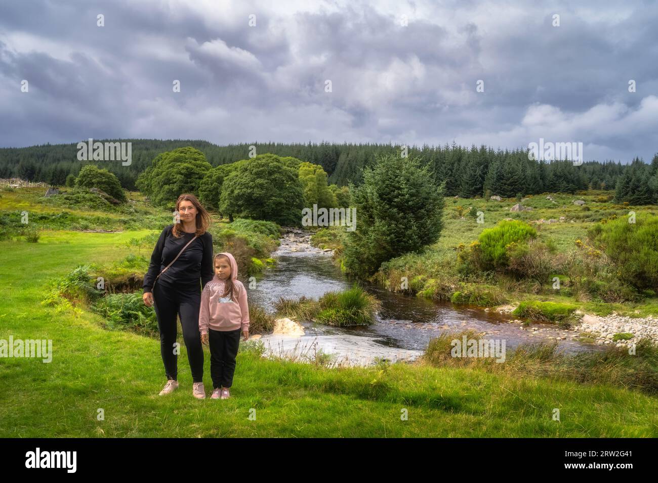 Mother and daughter standing on grass, with winding river and green ...