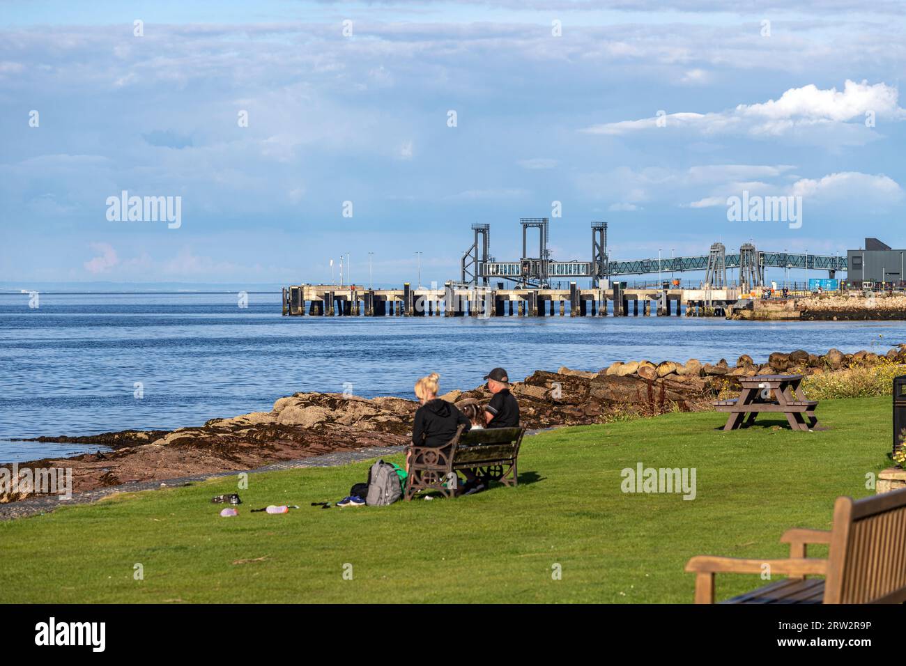 Couple in a wooden bech and the CalMac Ferries Brodick Ferry pier ...