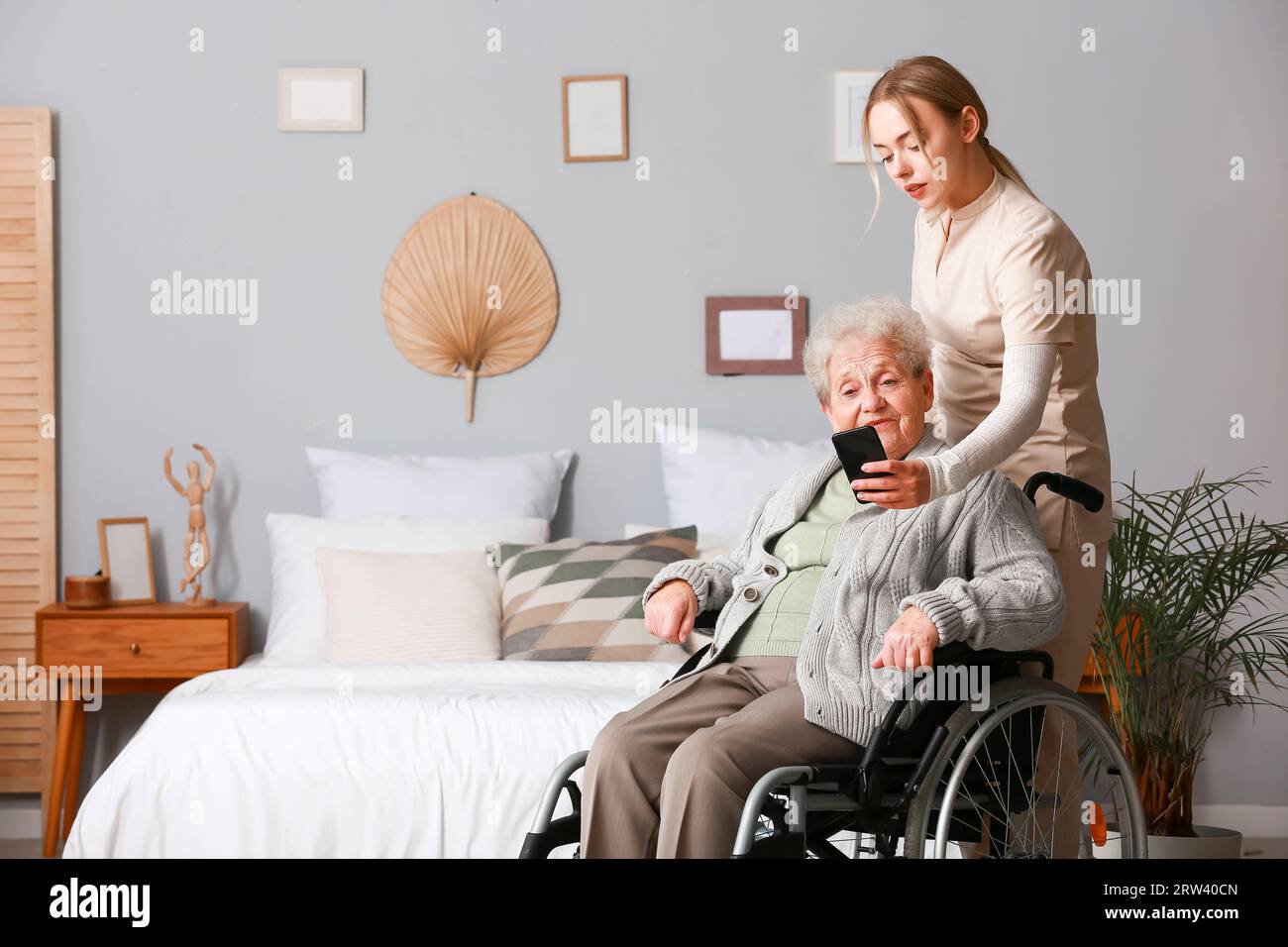 Young caregiver with senior woman in wheelchair using mobile phone at ...