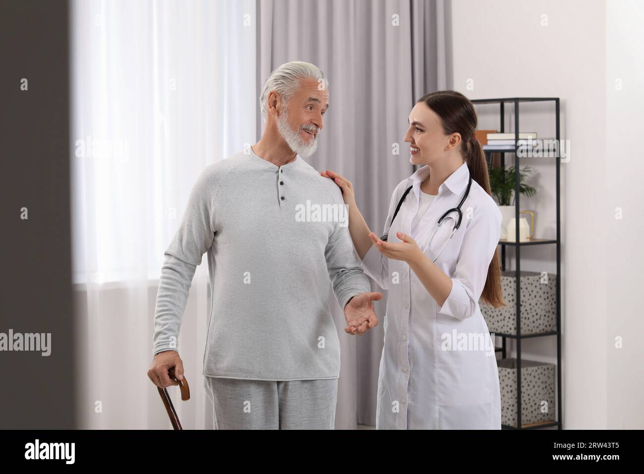 Young healthcare worker assisting senior man with walking cane indoors ...