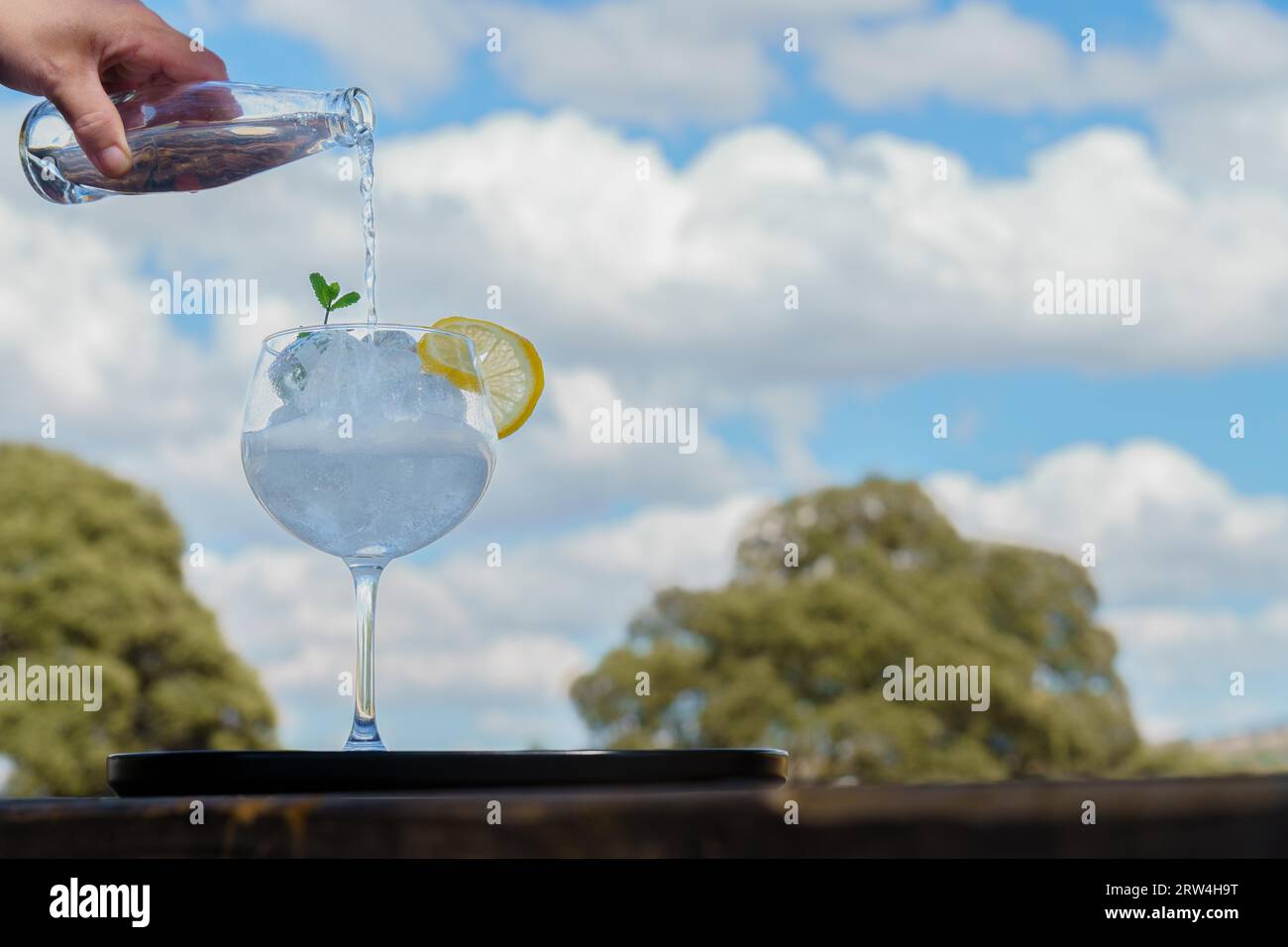 Woman's hand pouring tonic from a bottle into a glass with ice, preparing a gin and tonic in the ...