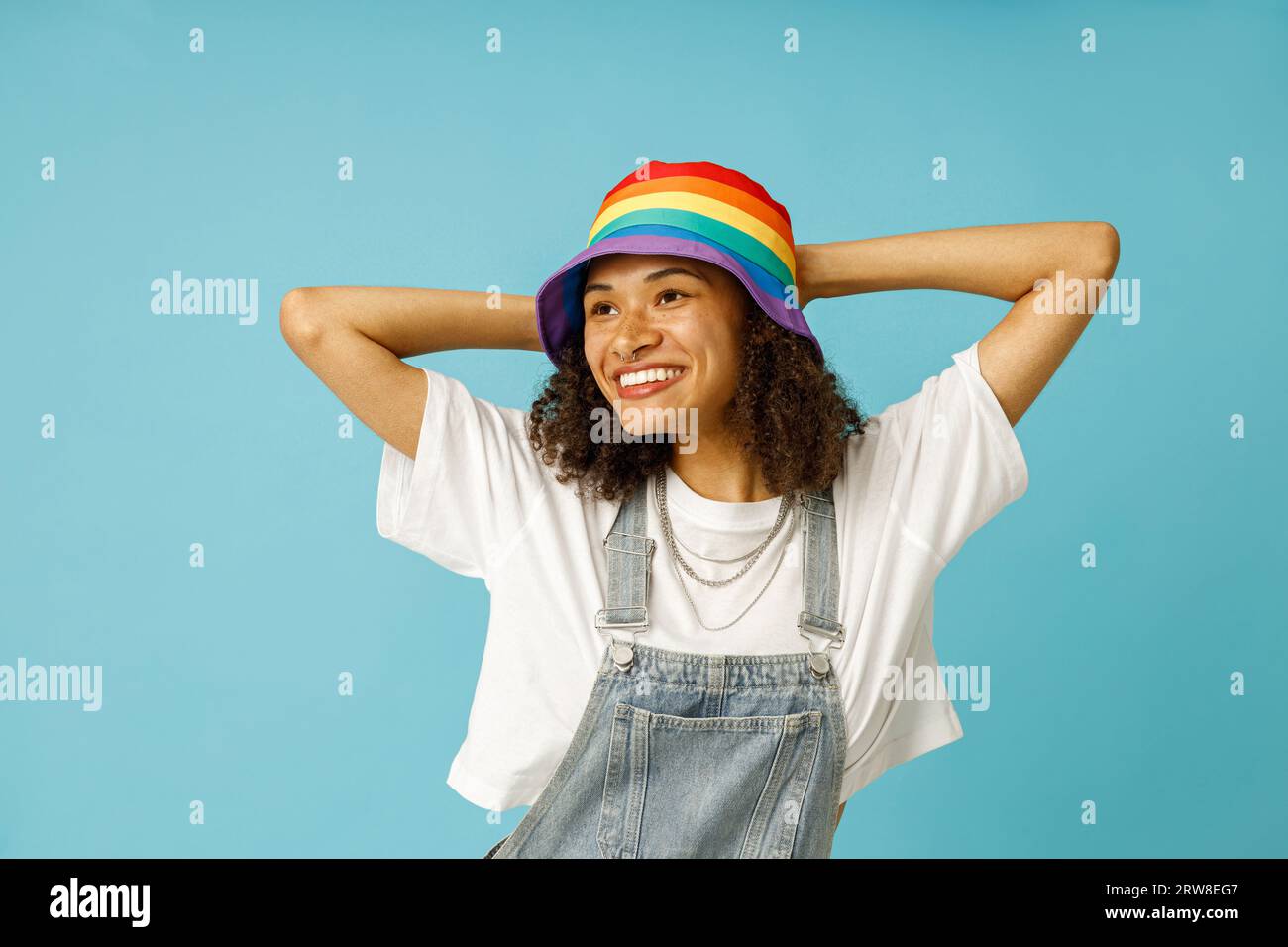 Cheerful woman in rainbow cap standing with raised hands and looks away on blue studio ...