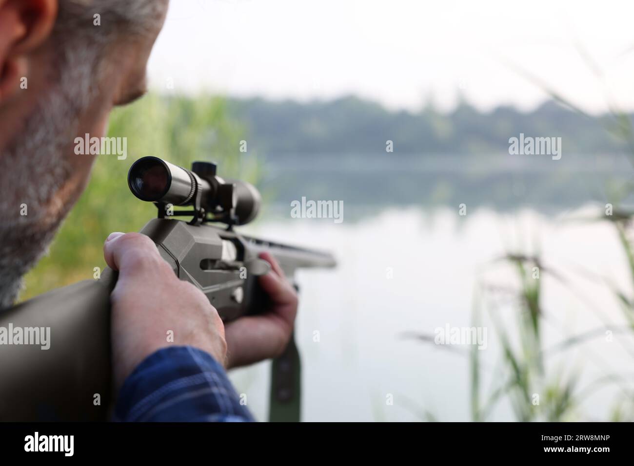 Man aiming with hunting rifle near lake outdoors, closeup. Space for ...