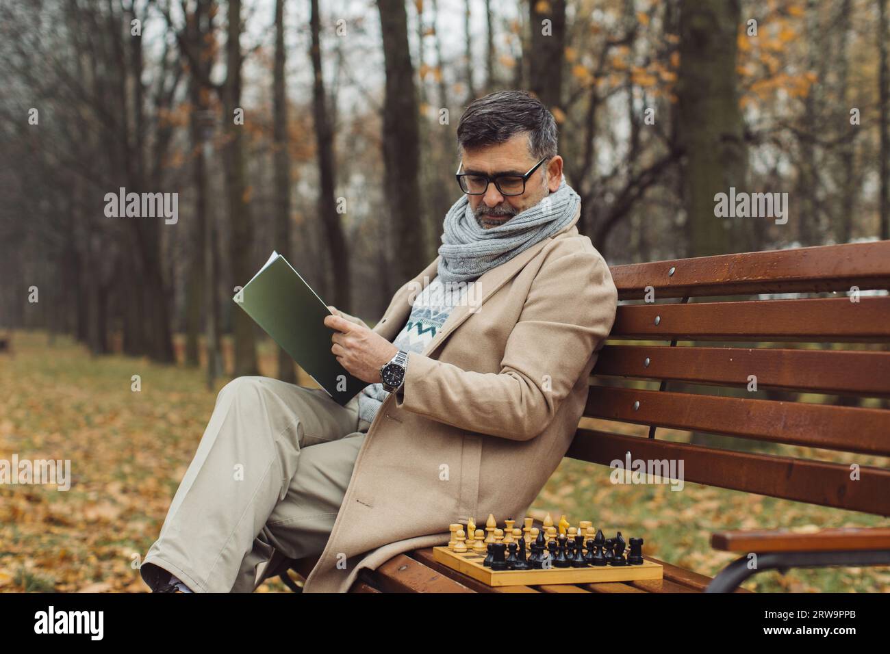 An elderly man reads a magazine while sitting on a bench in an autumn ...