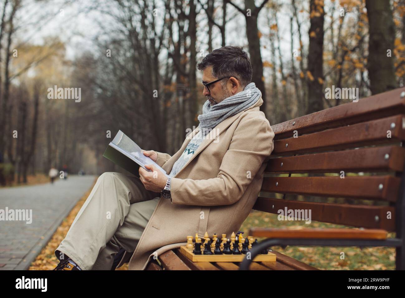 An elderly man reads a magazine while sitting on a bench in an autumn ...