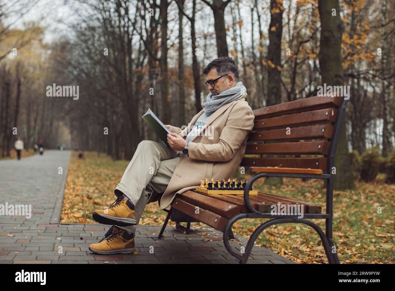 An elderly man reads a magazine while sitting on a bench in an autumn ...