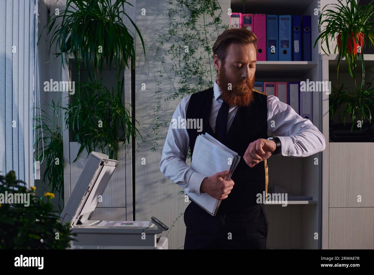 bearded businessman with papers looking at wristwatch near copier while ...
