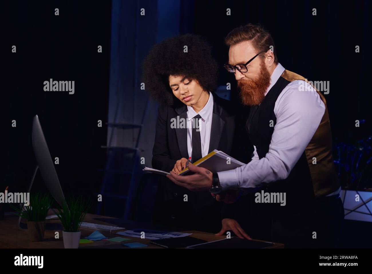 african american businesswoman and bearded colleague looking at documents in dark office, night ...