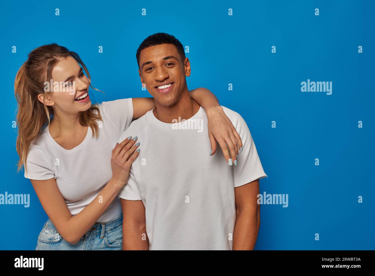 happy young woman embracing african american friend on blue backdrop ...