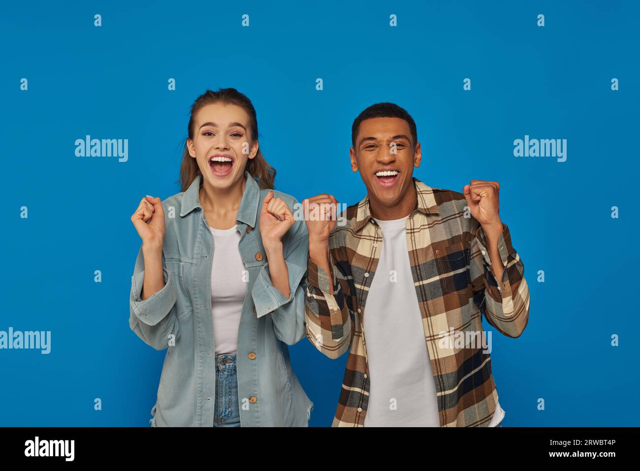 excited interracial couple screaming from joy looking at camera on blue ...