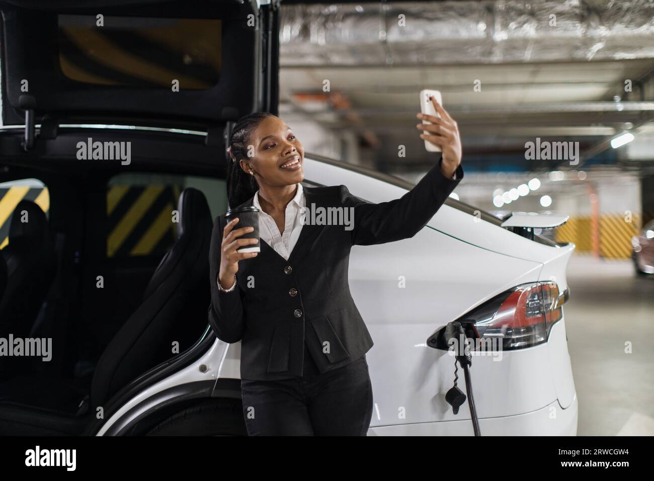 Lady with drink smiling at camera near charging EV in garage Stock ...