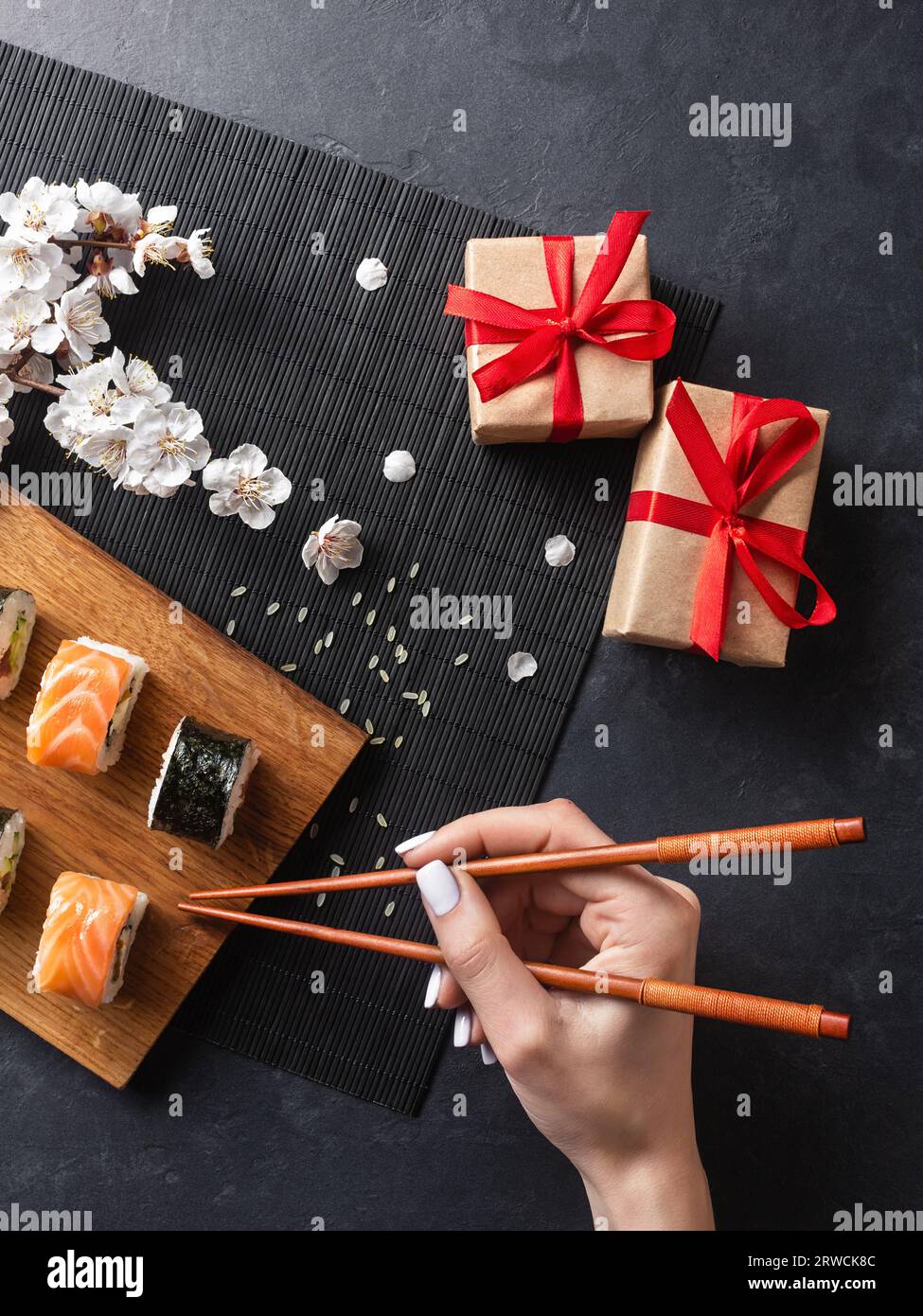 Set of sushi and maki rolls, hand with chopsticks, gift boxes and branch of white flowers on ...