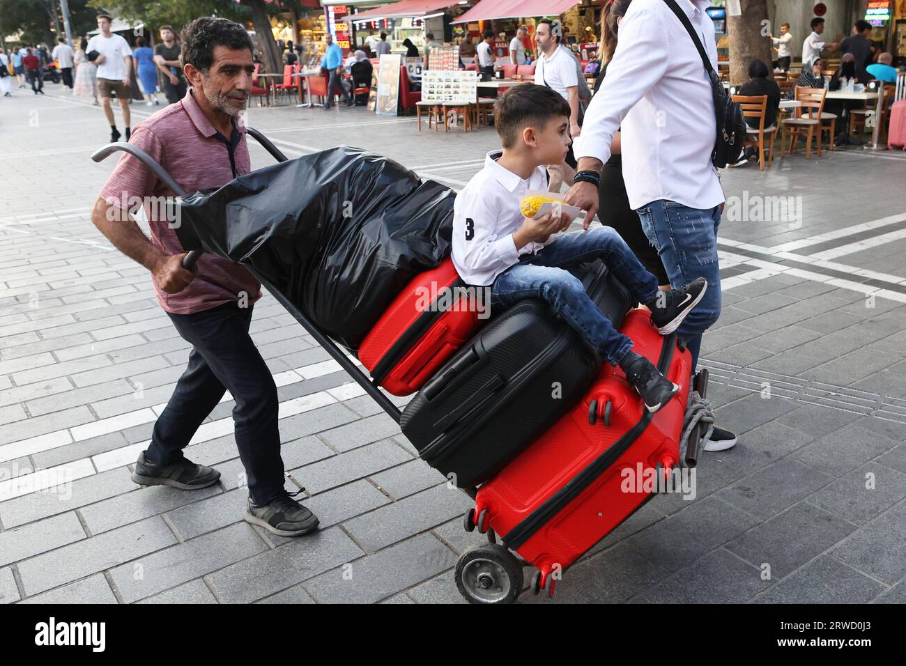 A father pushes a trolley filled with luggage with his son, eating corn ...