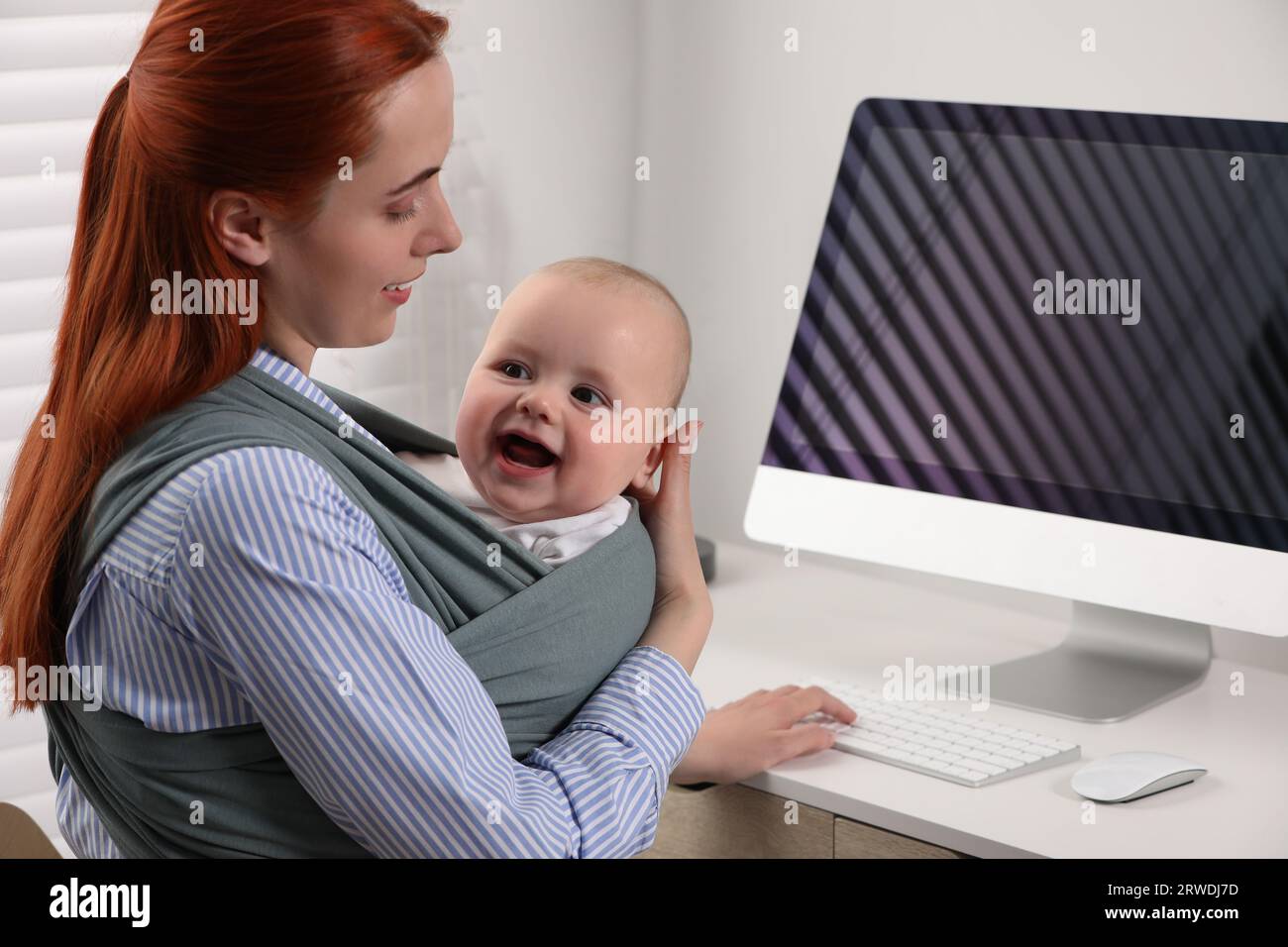 Mother holding her child in sling (baby carrier) while using computer ...