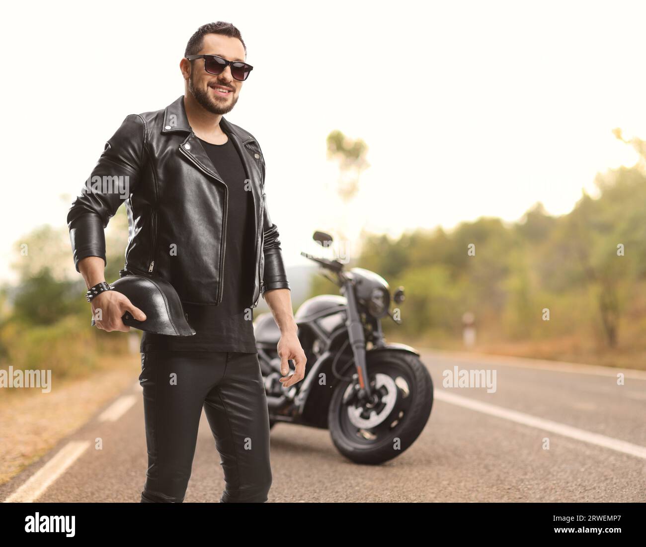 Biker in a leather jacket with parked motorcycle on the road holding helmet and wearing ...