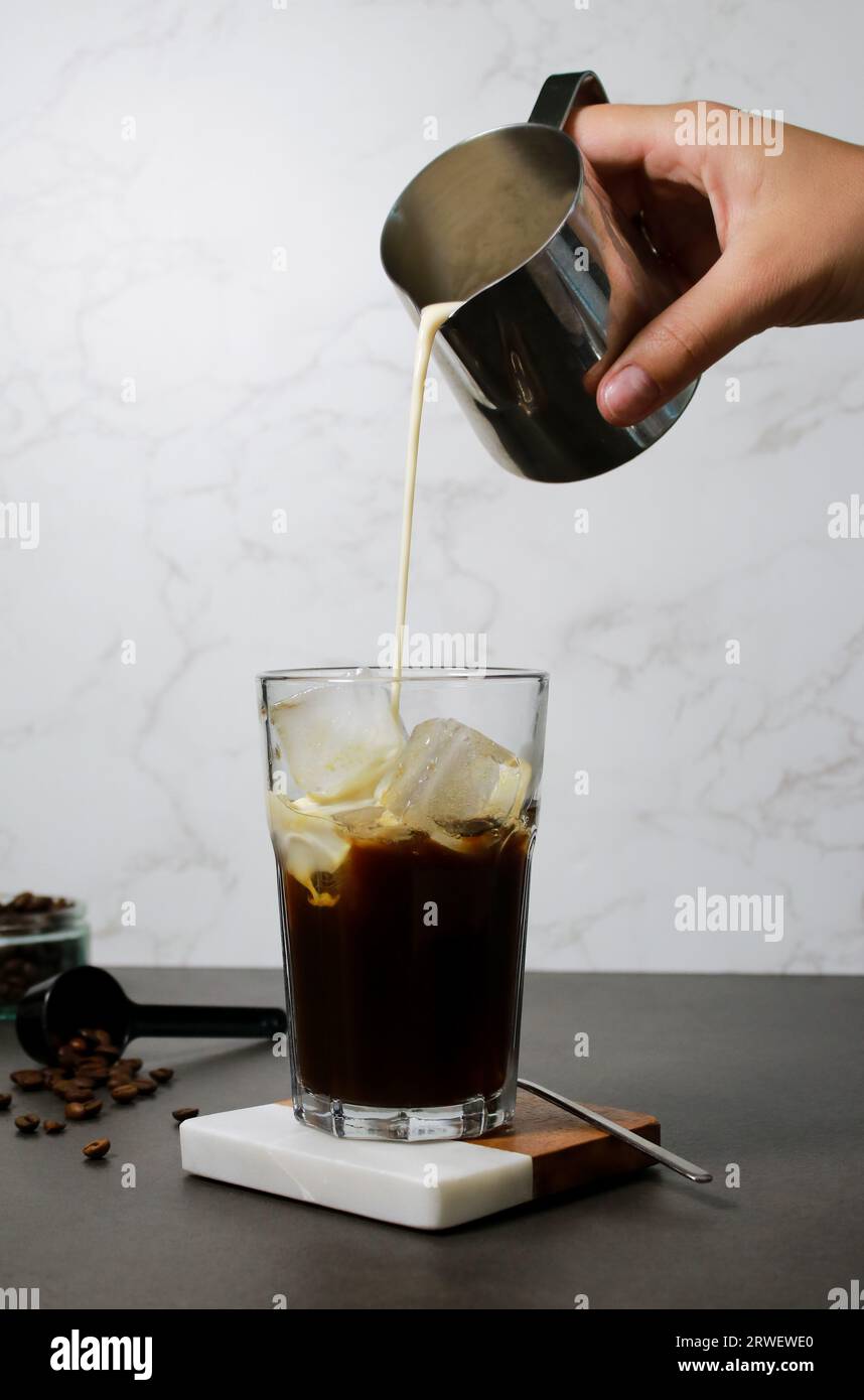 Pouring oat milk into cold ice coffee in a tall glass, grey marble ...