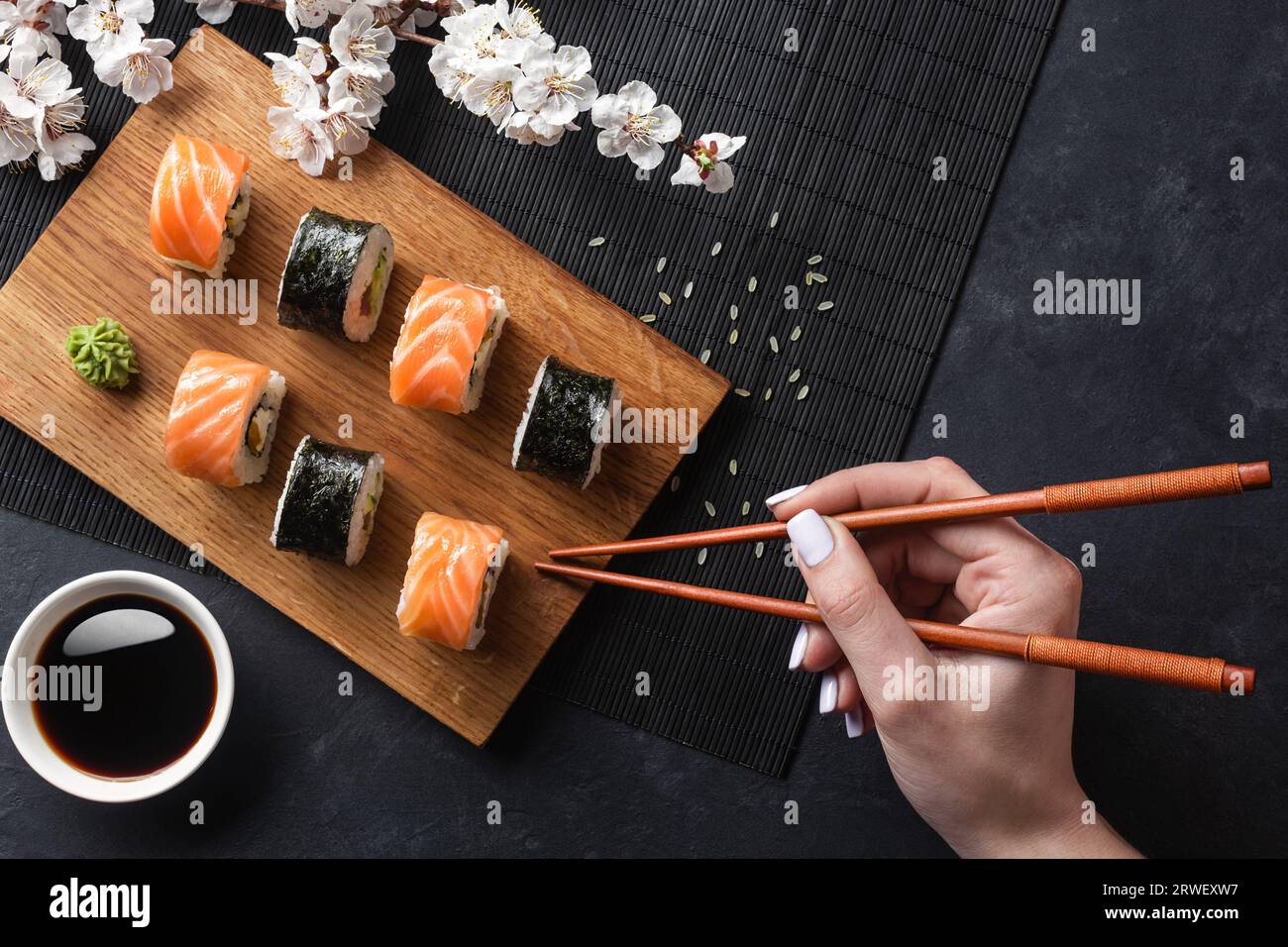 Set of sushi, maki rolls, hand with chopsticks and branch of white flowers on stone table. Top ...