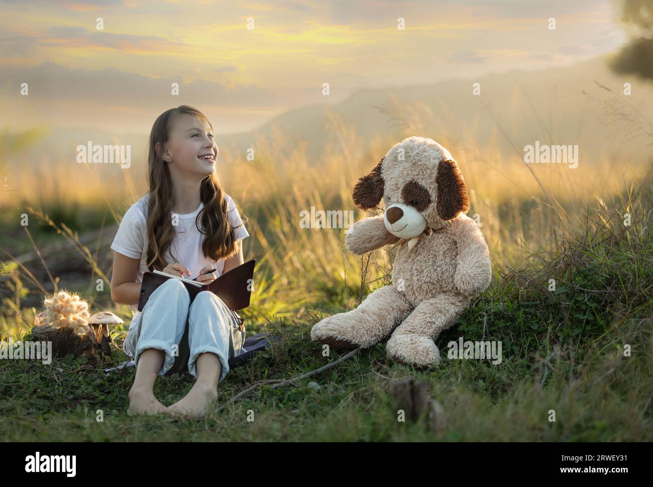 Young, smiling girl sitting in nature at sunset with her teddy writing ...