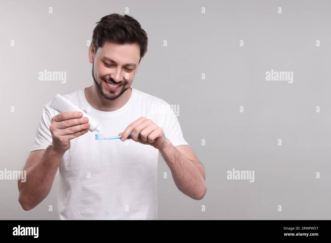 Happy man squeezing toothpaste from tube onto toothbrush on light grey ...