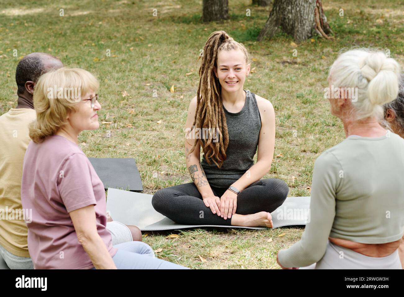 Young coach sitting on exercise mat and talking to senior people before ...