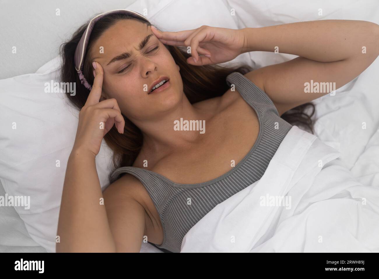 A lady in grimaces, temples aching, hands cradling her head, battling a headache on a bright bed ...