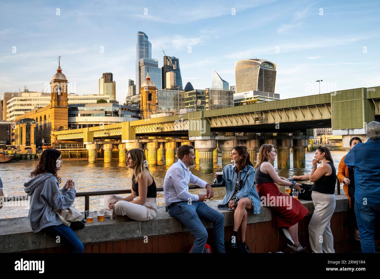 Young People Sitting By The River Thames In The Early Evening Drinking ...